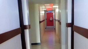 Um corredor de hotel com paredes brancas, detalhes em castanho e uma porta vermelha no final.