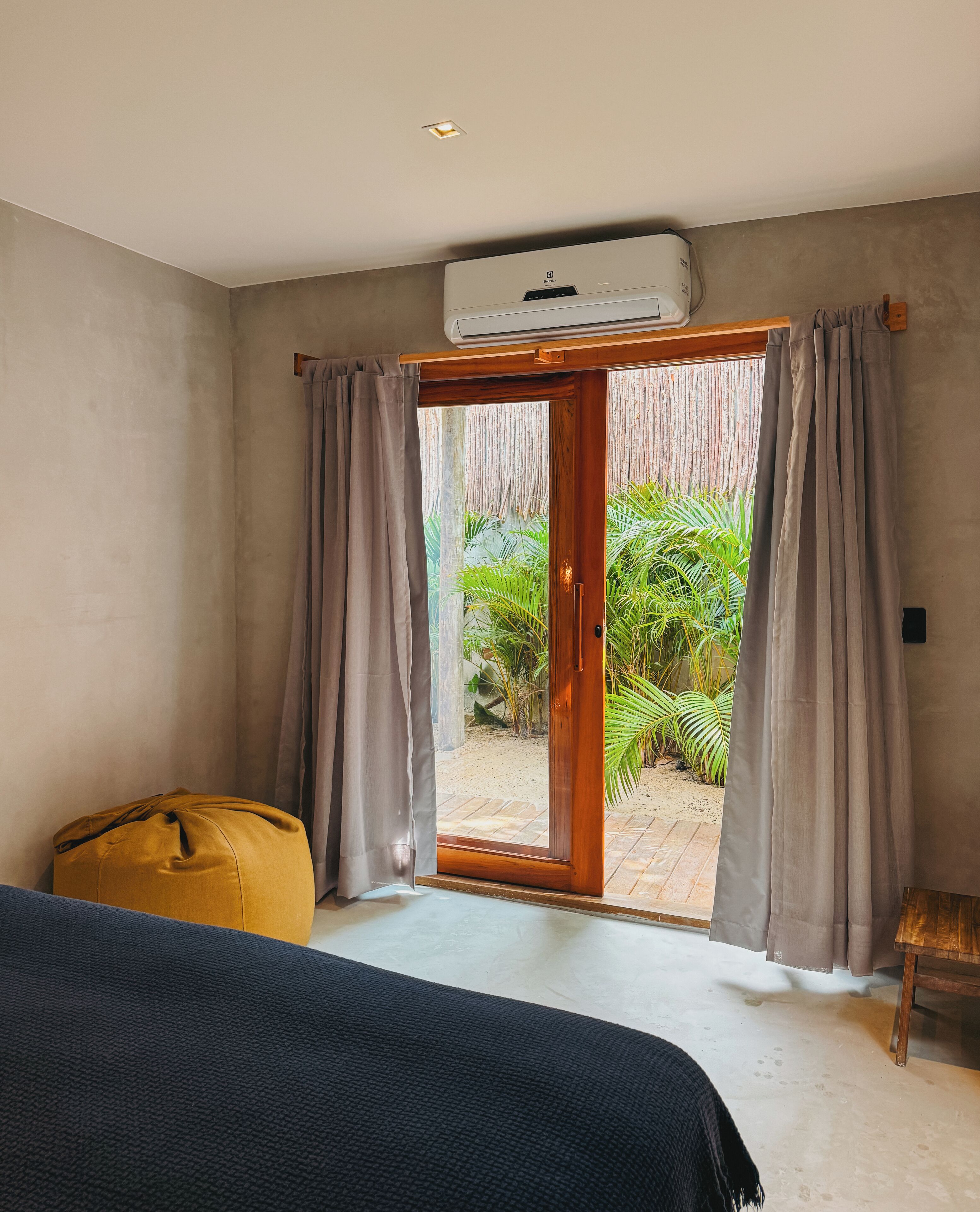 Quarto com cama, um pequeno banco de madeira, um banquinho amarelo e uma porta de correr de vidro que dá para uma área externa com plantas.