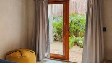 Quarto com cama, um pequeno banco de madeira, um banquinho amarelo e uma porta de correr de vidro que dá para uma área externa com plantas.