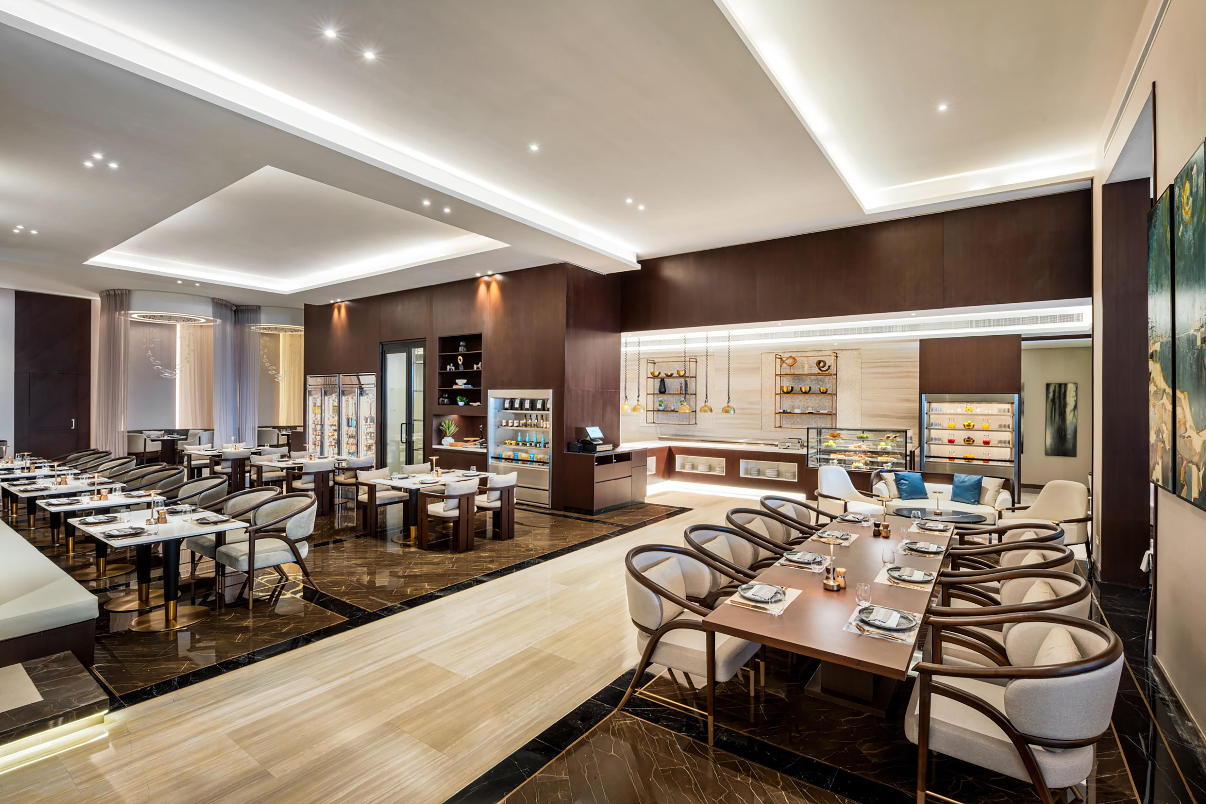 Espaço de refeições moderno em hotel, com várias mesas e cadeiras, área de bar central e vista para a paisagem urbana.