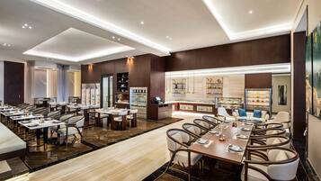 Espaço de refeições moderno em hotel, com várias mesas e cadeiras, área de bar central e vista para a paisagem urbana.