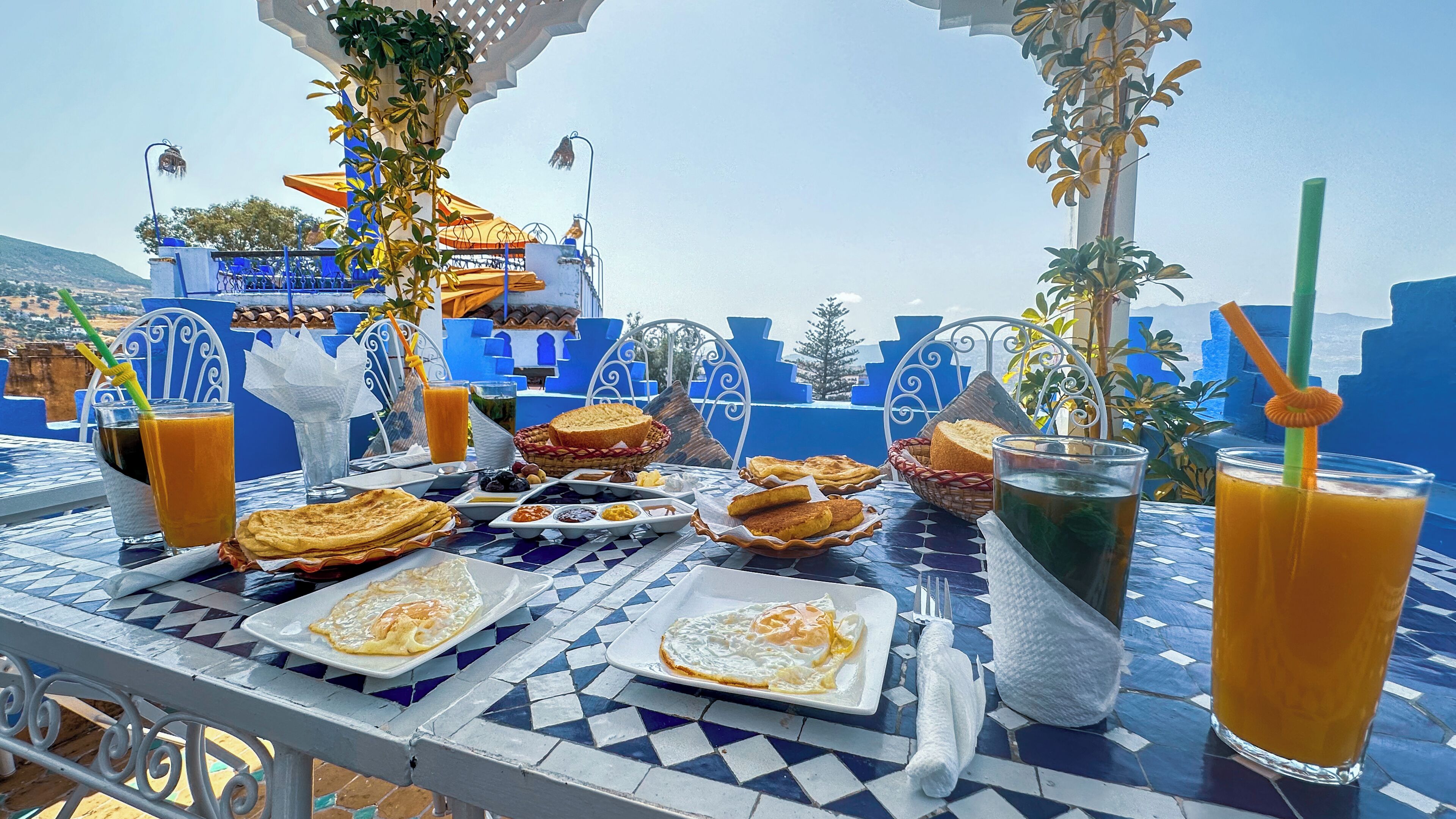 Mesa posta com itens de café da manhã, incluindo ovos, torradas e bebidas, com vista para um edifício azul ao fundo.
