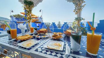 Mesa posta com itens de café da manhã, incluindo ovos, torradas e bebidas, com vista para um edifício azul ao fundo.