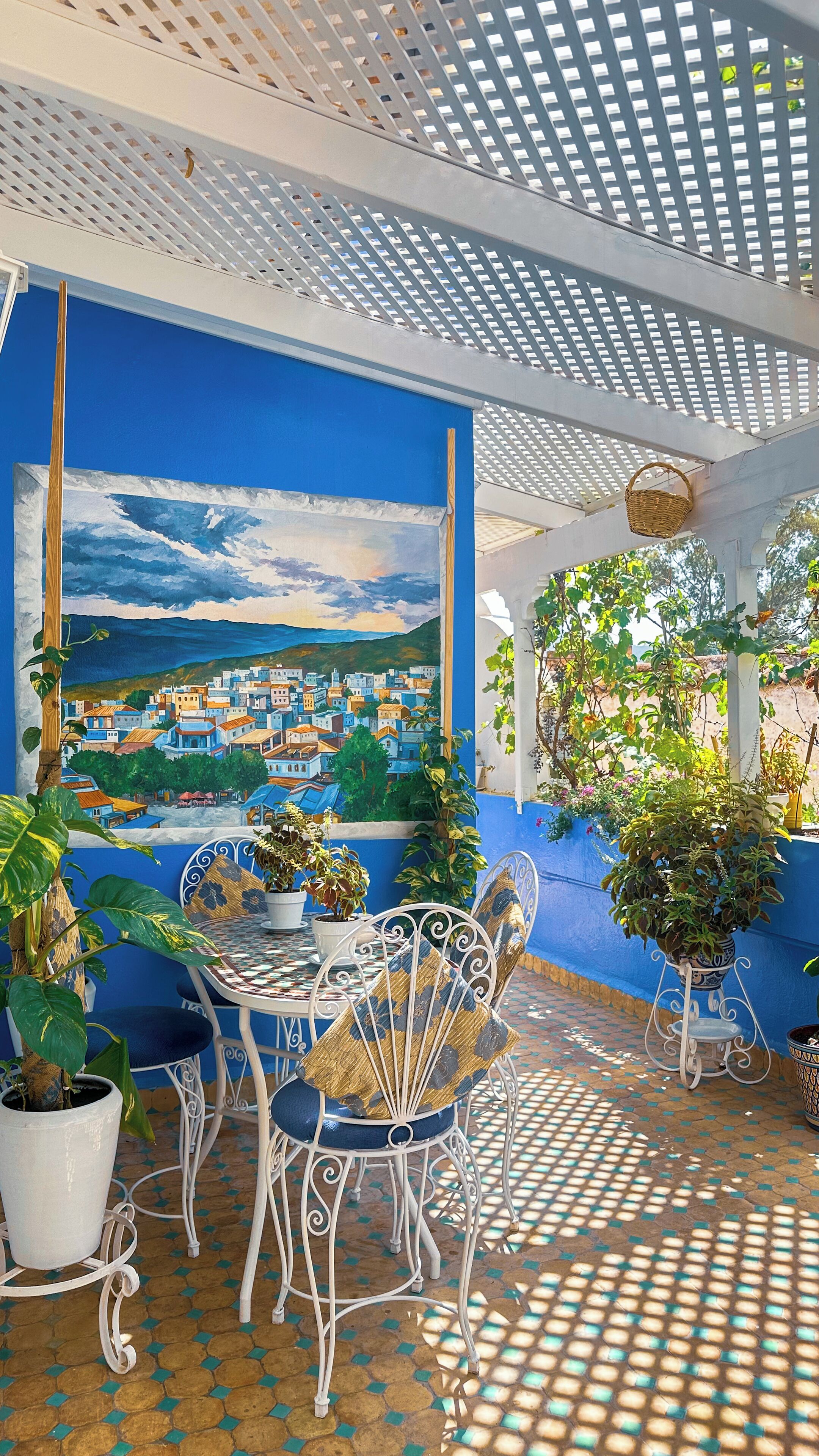 Um alpendre coberto com um mural azul, móveis de metal branco e plantas em vasos.