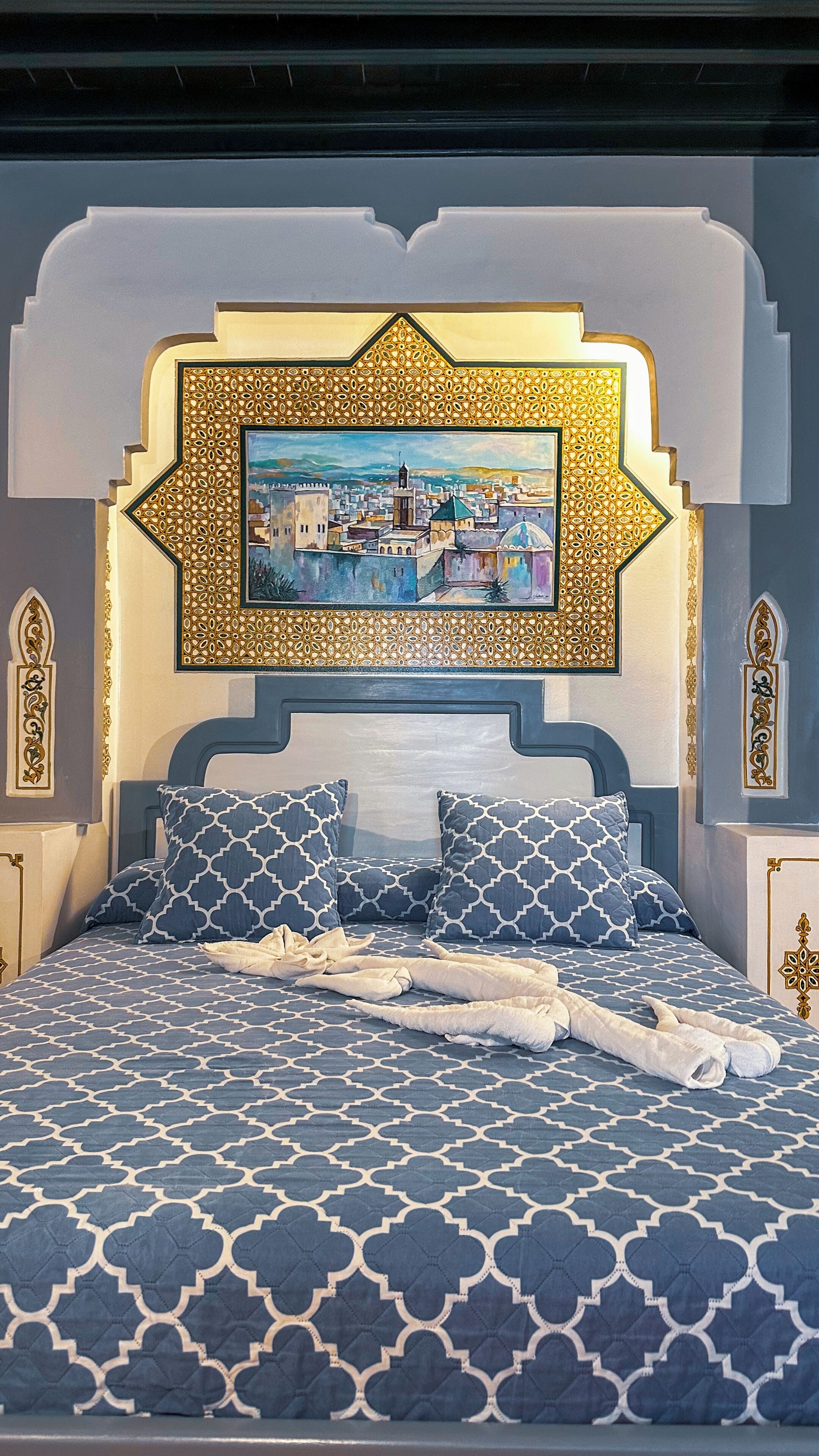 Uma cama com colcha estampada em azul e branco, uma pintura de paisagem urbana acima e painéis decorativos na parede.