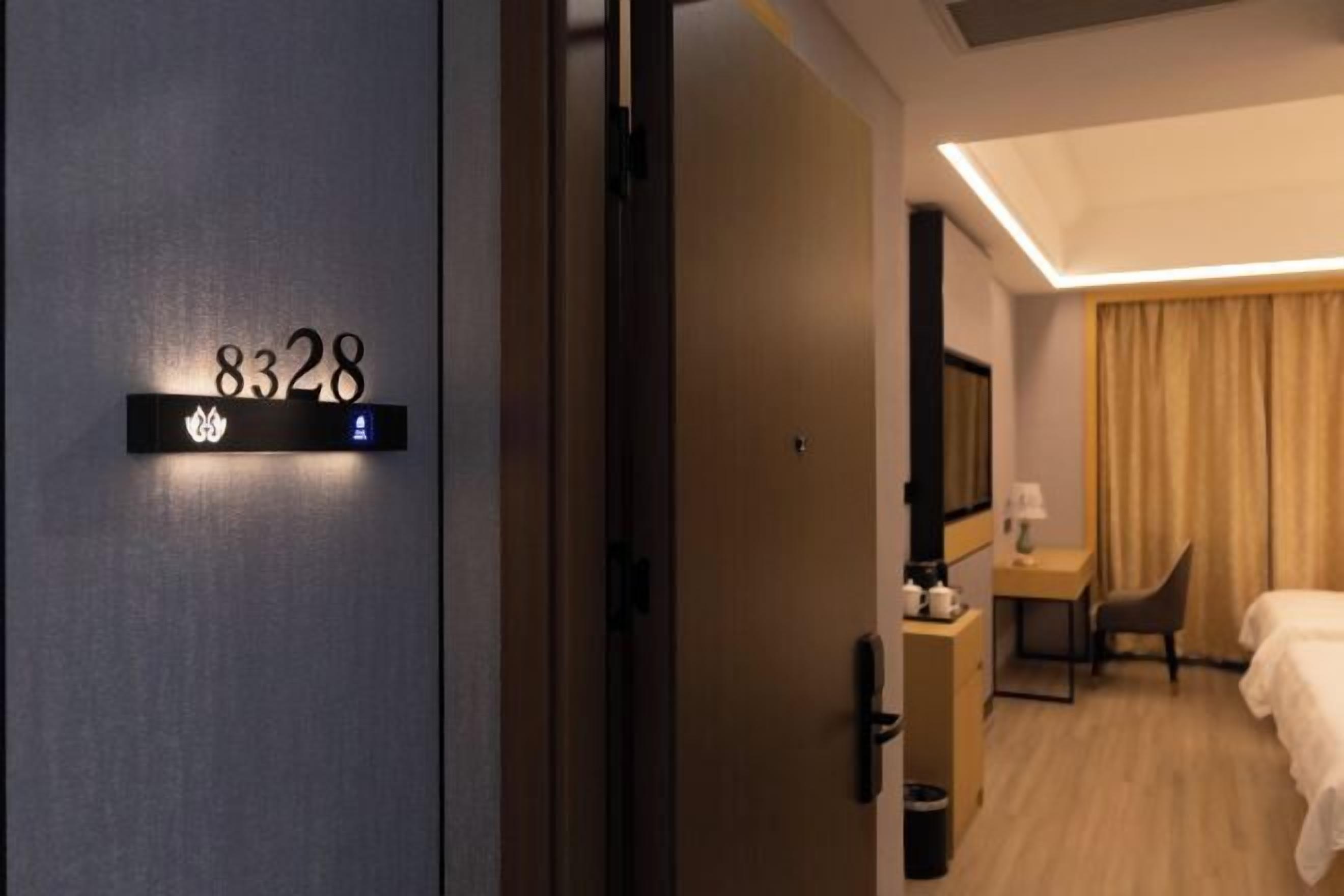 Quarto de hotel com placa de número, uma escrivaninha com luminária, uma cadeira e uma cama.