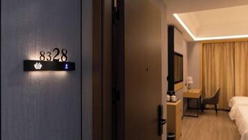 Quarto de hotel com placa de número, uma escrivaninha com luminária, uma cadeira e uma cama.