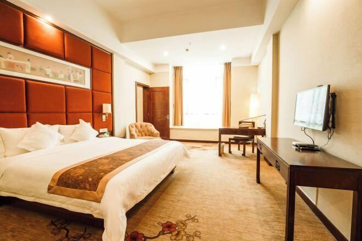 Quarto de hotel com uma cama grande, uma escrivaninha, uma cadeira, uma televisão e uma janela com cortinas.