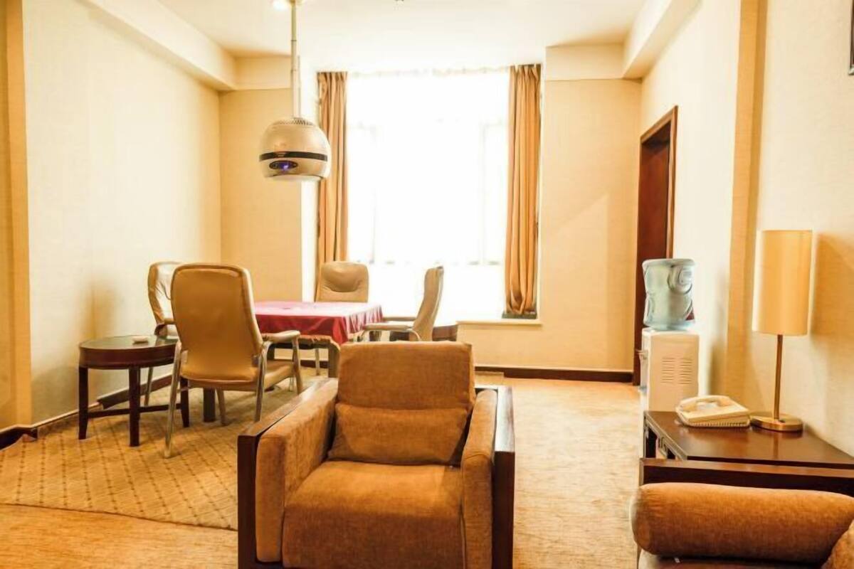 Quarto de hotel com sofá, mesa de jantar com cadeiras, luminária e janela com cortinas.