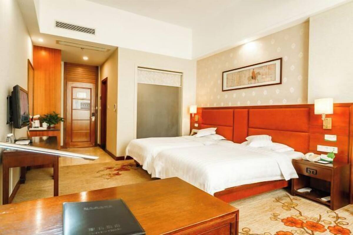Quarto de hotel com uma cama de casal, uma escrivaninha com cadeira, mesa de cabeceira, televisão e uma luminária na parede.