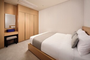 Um quarto com cama, um banquinho, cômoda, espelho e guarda-roupa.