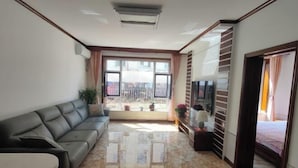 Uma sala de estar com sofá, mesa pequena, televisão e uma janela ampla com cortinas.