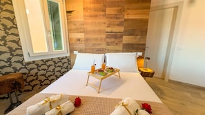 Uma cama bem arrumada com lençóis brancos, uma bandeja com frutas e bebidas, e uma rosa vermelha sobre a cama.