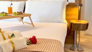 Uma cama bem arrumada com lençóis brancos, uma mesinha redonda com uma travessa de frutas, uma rosa vermelha e um abajur sobre um criado-mudo de madeira.