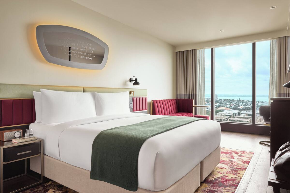 Quarto de hotel com uma cama grande, uma mesa de cabeceira, uma cadeira vermelha, uma escrivaninha e uma janela ampla com vista para a cidade.