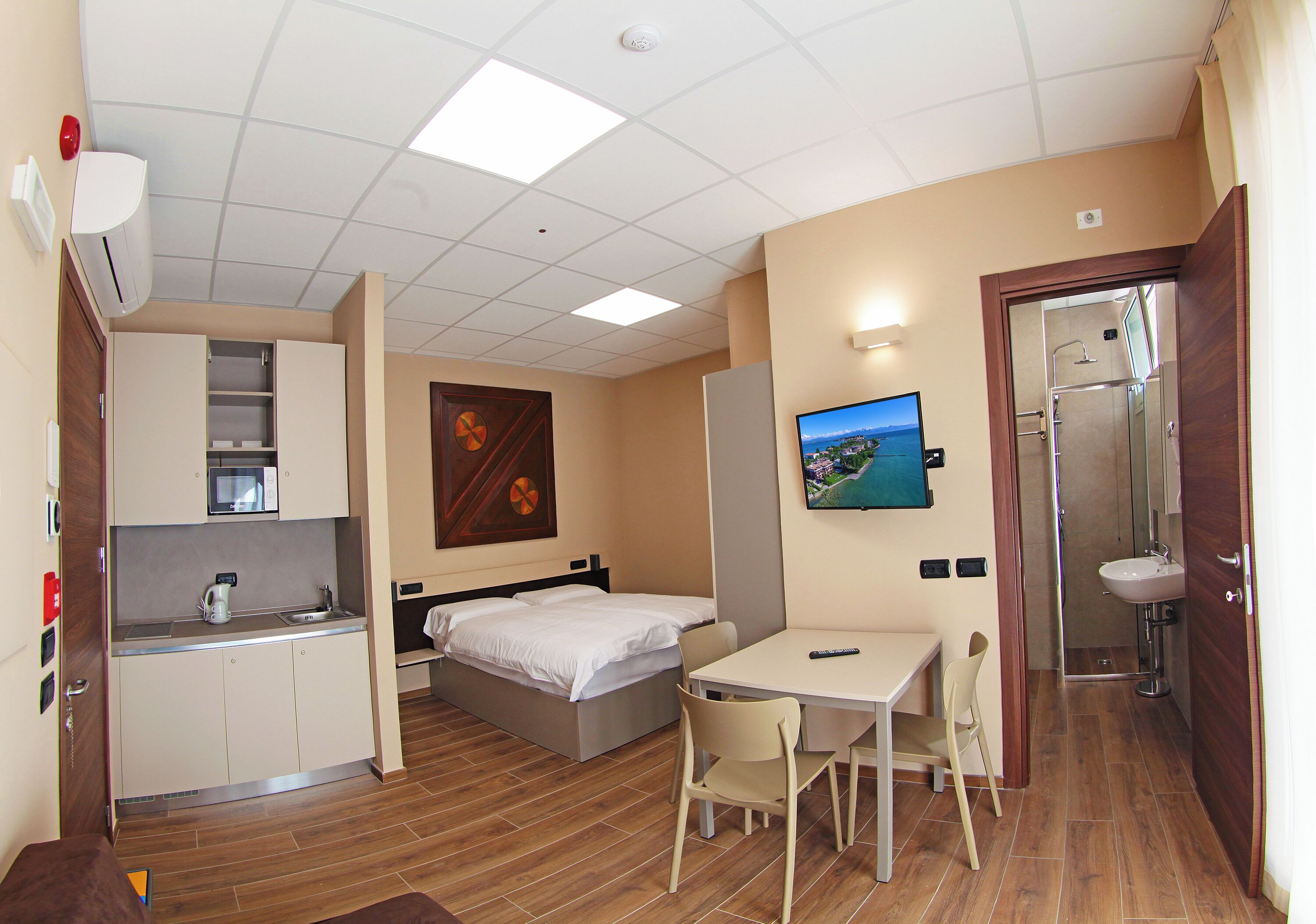 Quarto de hotel compacto com cozinha compacta, uma cama, uma mesa pequena com cadeiras, televisão e banheiro.