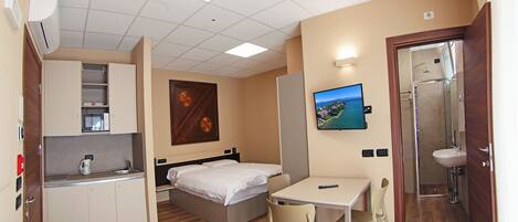 Quarto de hotel compacto com cozinha compacta, uma cama, uma mesa pequena com cadeiras, televisão e banheiro.