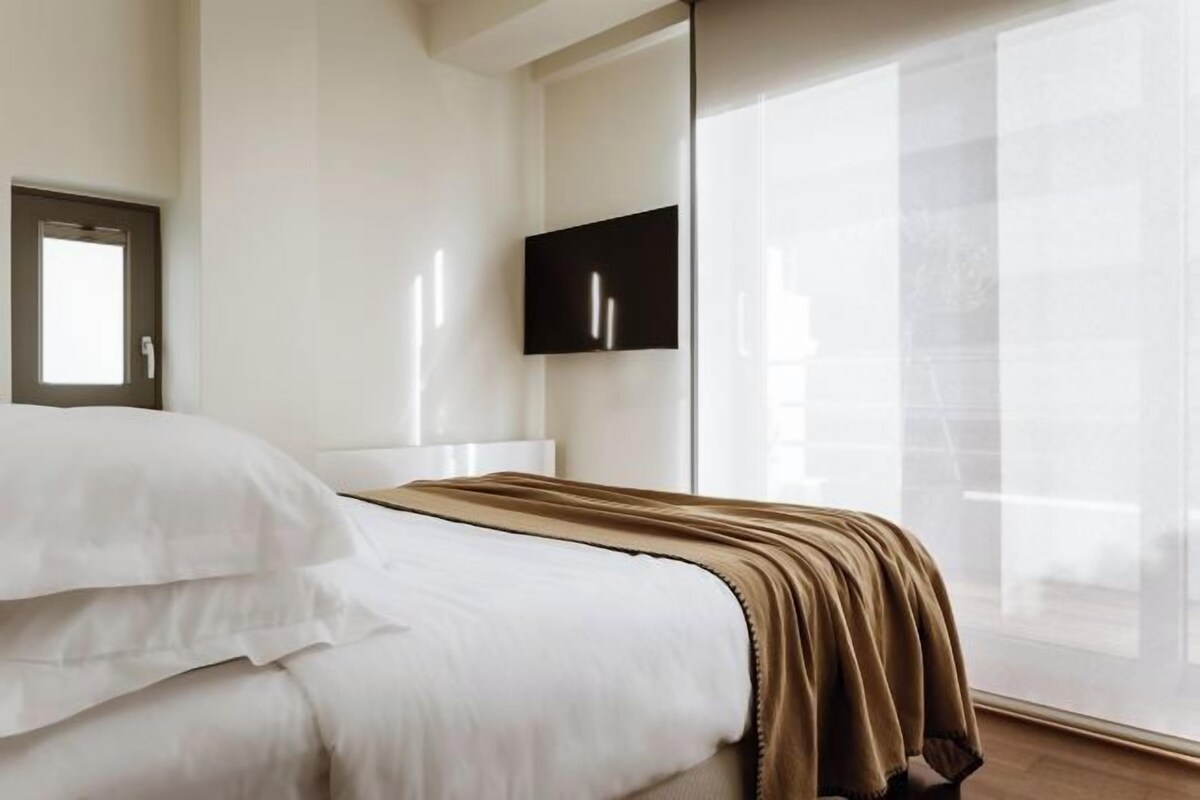 Quarto de hotel com uma cama, televisão na parede e uma janela grande.