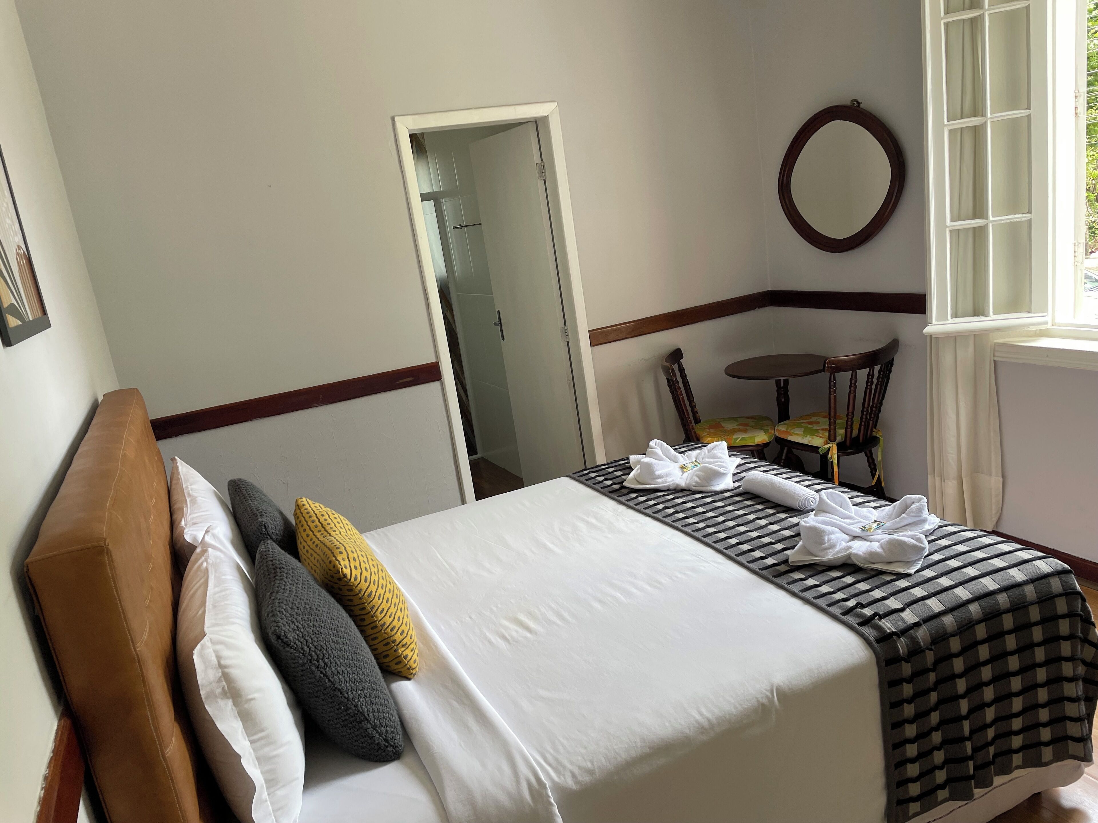 Cama bem arrumada com manta xadrez, cabeceira de madeira e várias almofadas. Há uma mesa redonda pequena com cadeiras e um espelho na parede.