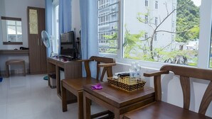 Um quarto com escrivaninha de madeira, televisão, janela com vista para edifícios e árvores, um ventilador e uma pequena mesa com uma cesta.