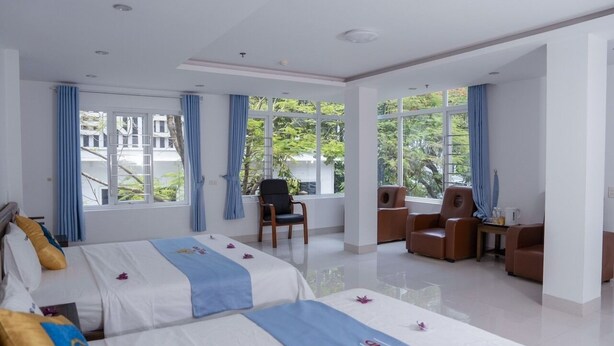 Quarto de hotel com duas camas, área de estar com cadeiras e janelas amplas com vista para o verde.