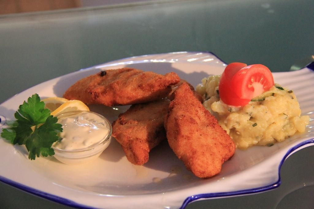Um prato com dois filés de peixe empanados, purê de batatas, uma fatia de tomate, um gomo de limão e uma pequena tigela de molho.