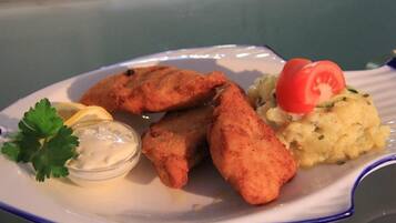 Um prato com dois filés de peixe empanados, purê de batatas, uma fatia de tomate, um gomo de limão e uma pequena tigela de molho.