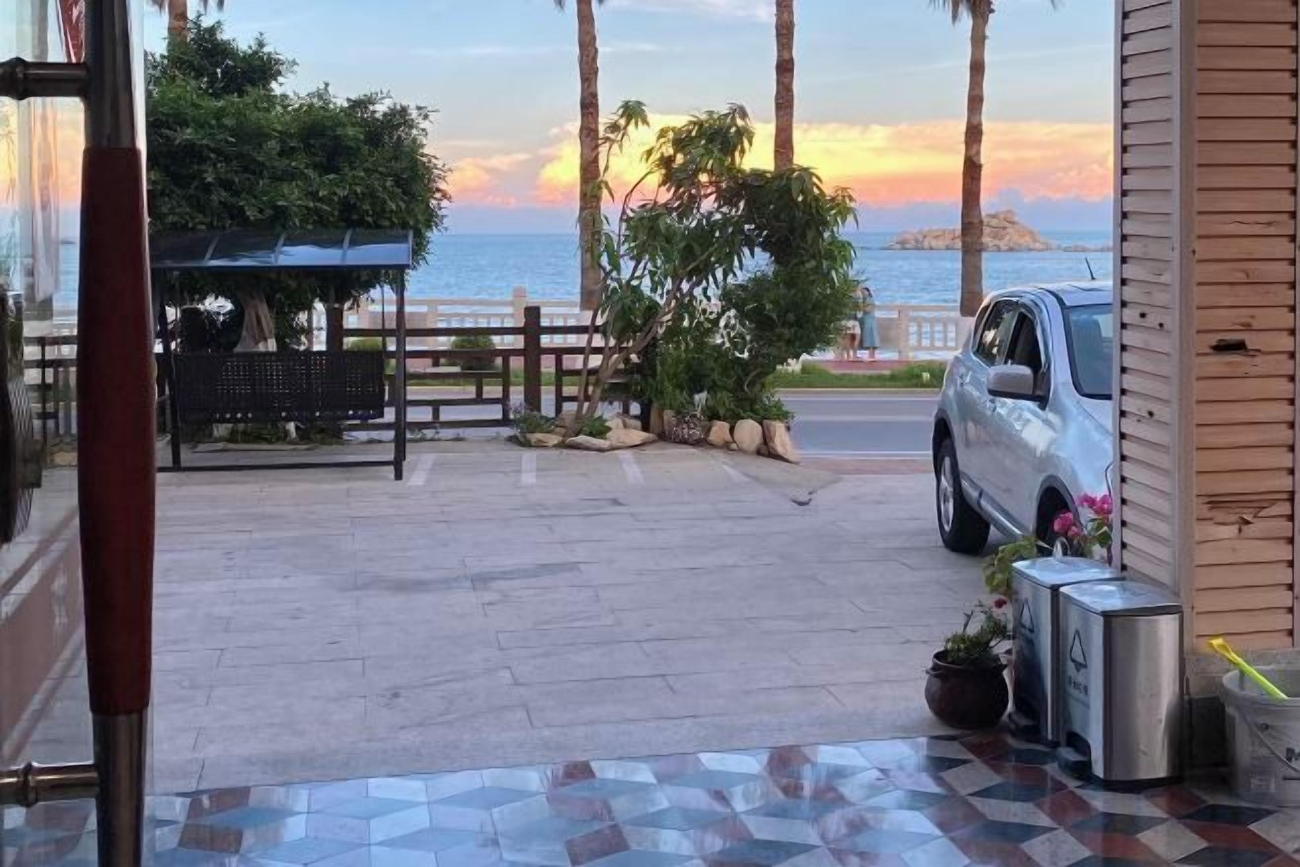 Um pátio pavimentado com um carro estacionado à direita, um banco e uma pequena construção com telhado.