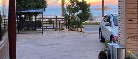 Um pátio pavimentado com um carro estacionado à direita, um banco e uma pequena construção com telhado.