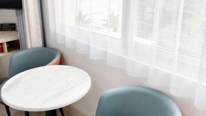 Quarto com uma mesa redonda de mármore, duas cadeiras verde-água, uma televisão e cortinas leves.