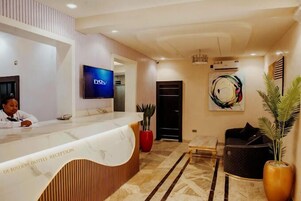 Uma área de recepção de hotel com balcão de mármore, televisão fixada na parede e mesa de atendimento.