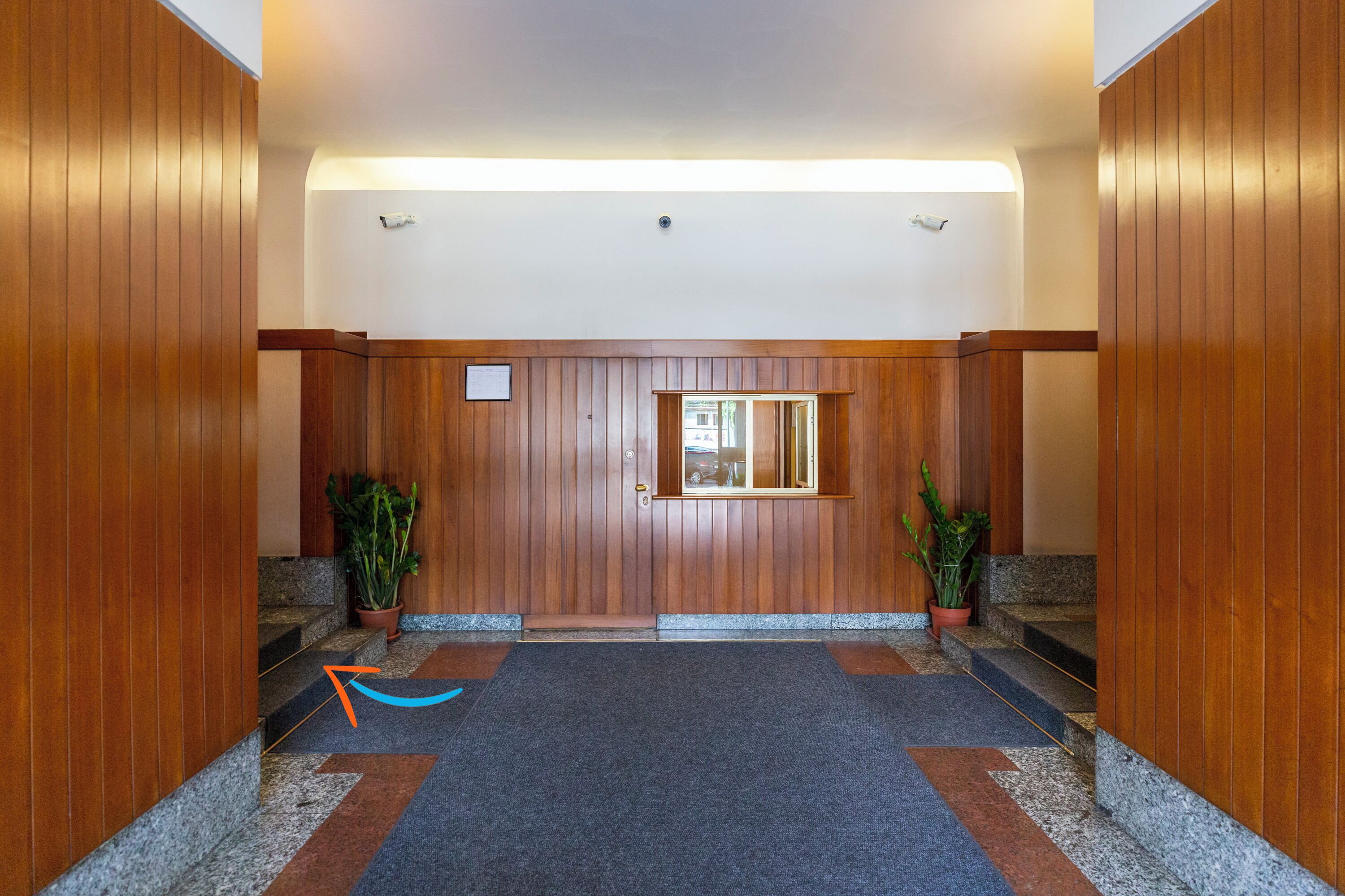 Um saguão de hotel com porta de madeira, plantas em vasos e piso coberto de carpete.