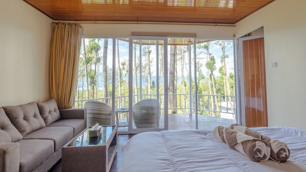 Quarto com uma cama grande, um sofá, uma mesinha e vista para árvores através de uma porta de vidro.