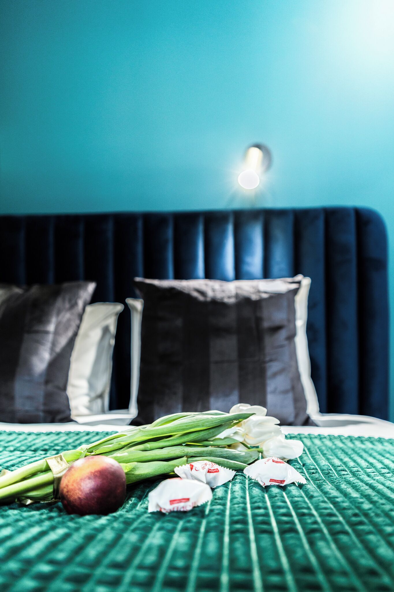 Uma cama com um edredom verde texturizado, um buquê de flores brancas e vários chocolates embalados sobre a cama.