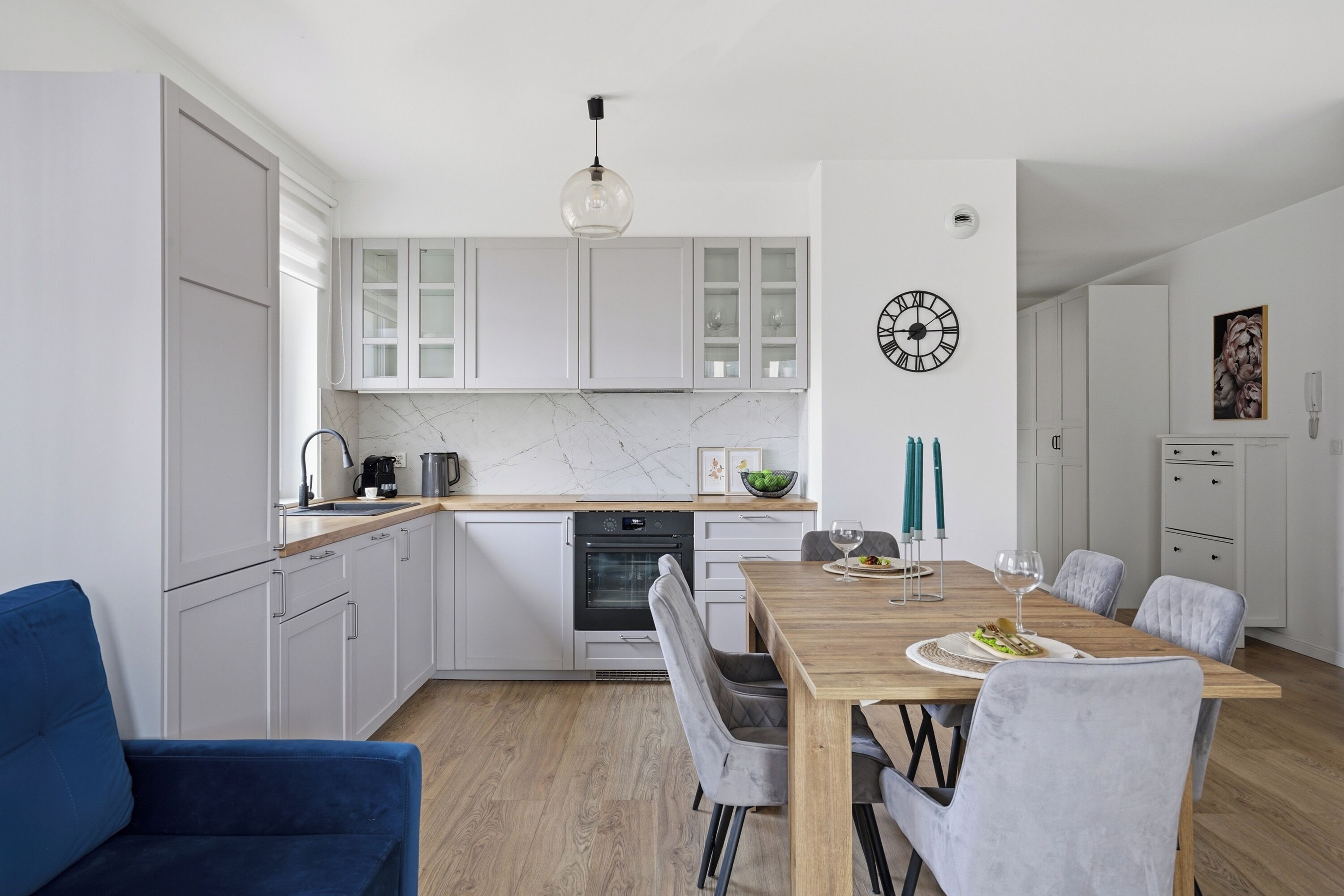 Cozinha moderna com armários brancos, bancada de mármore e área de refeições com mesa e cadeiras de madeira.