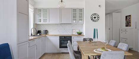 Cozinha moderna com armários brancos, bancada de mármore e área de refeições com mesa e cadeiras de madeira.