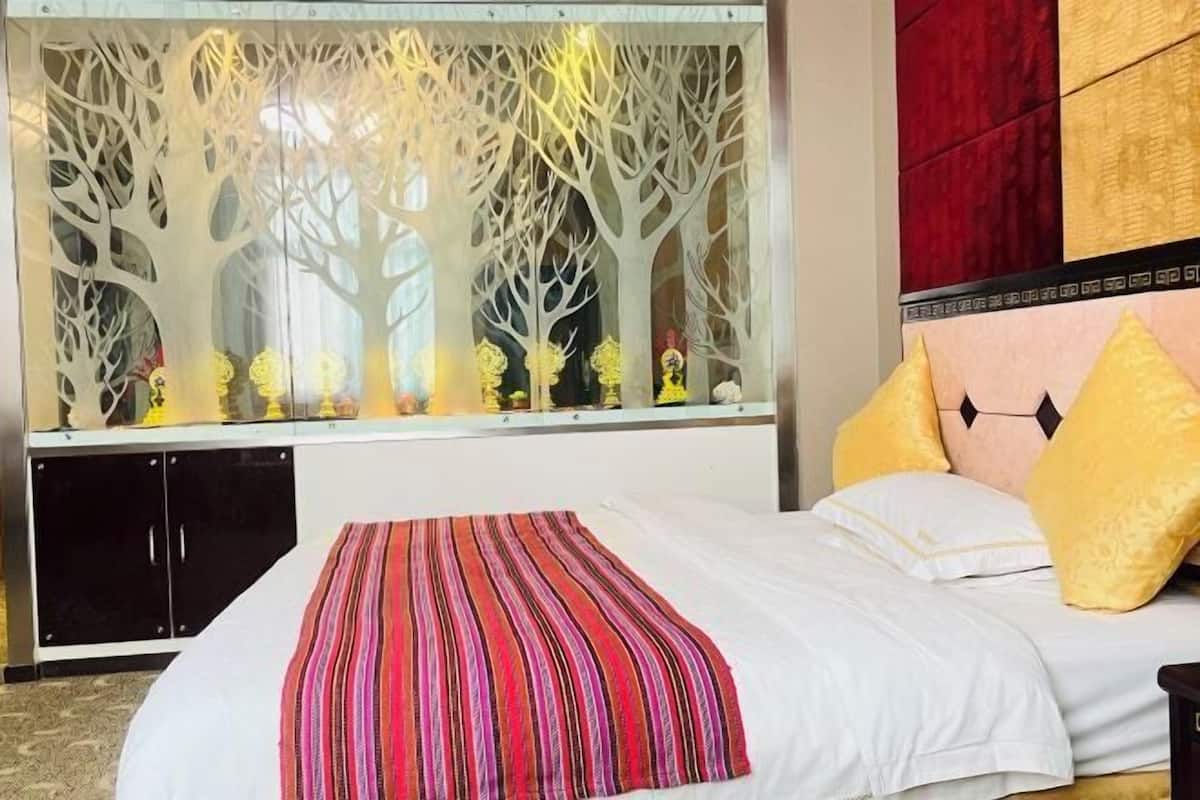 Quarto de hotel com cama, manta listrada, janela decorativa com desenho de árvore e cabeceira vermelha e dourada.