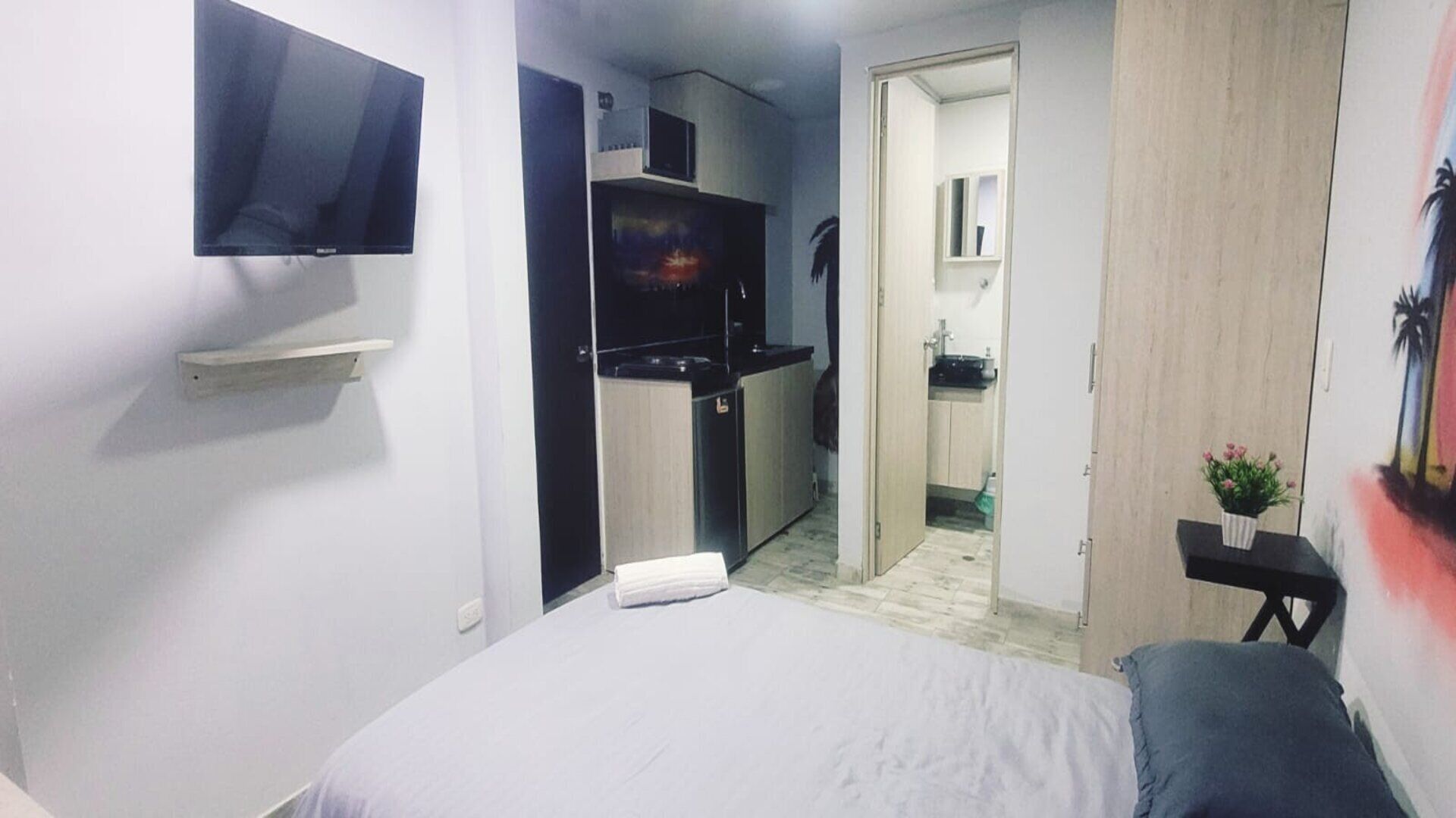 Quarto compacto com cama, secretária pequena, TV e kitchenette.