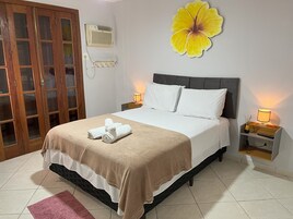 Cama bem arrumada com manta marrom, travesseiros brancos e toalha enrolada. Há um ar condicionado fixado na parede, uma porta de madeira com painéis de vidro e uma decoração de flor amarela.