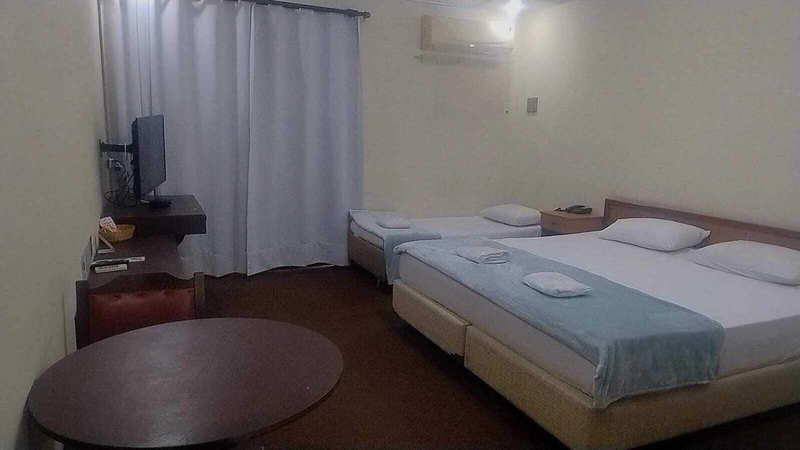 Quarto de hotel com duas camas, uma pequena mesa, televisão e janela com cortinas.