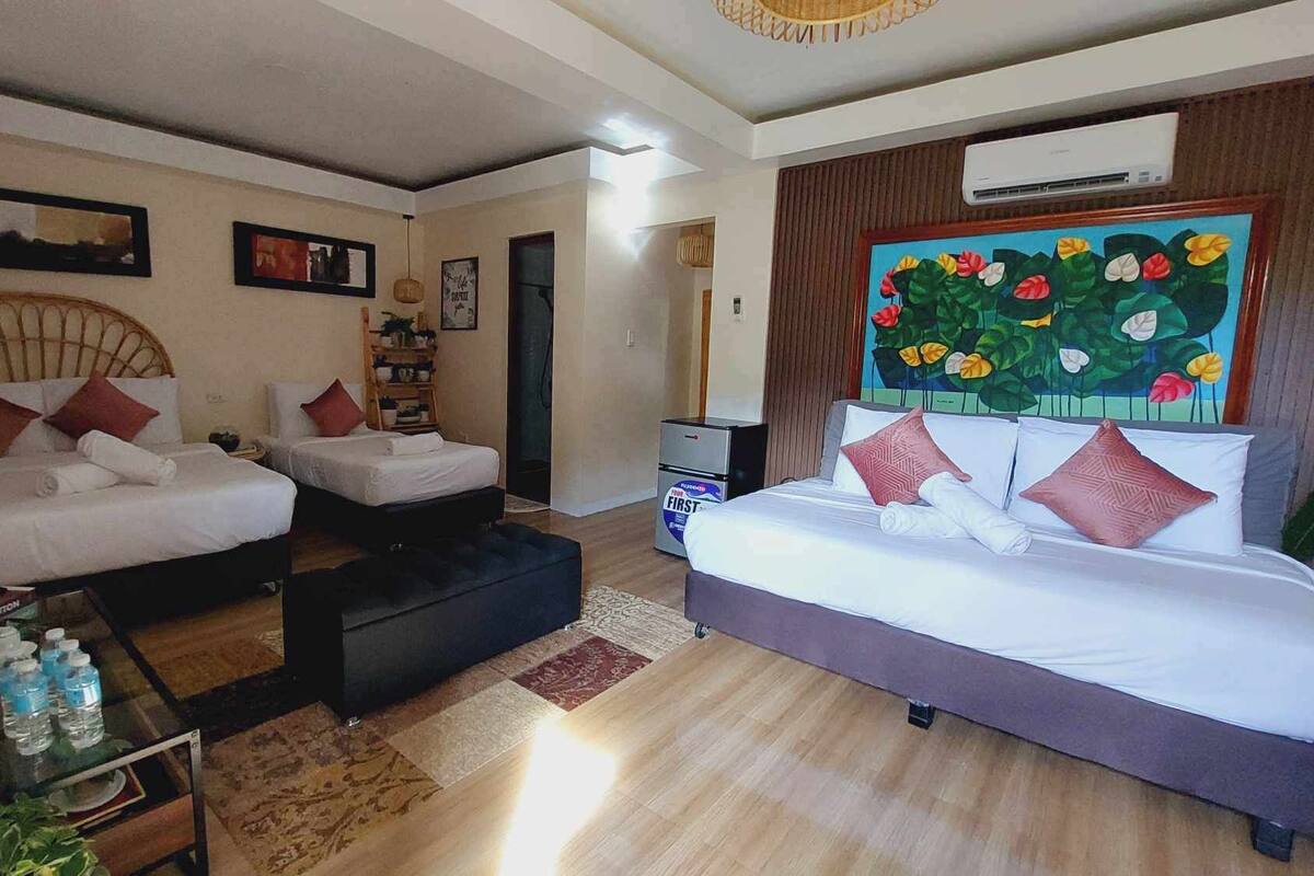 Quarto de hotel com duas camas de solteiro, uma cama de casal, um pequeno frigorífico, uma televisão e um quadro com flores na parede.