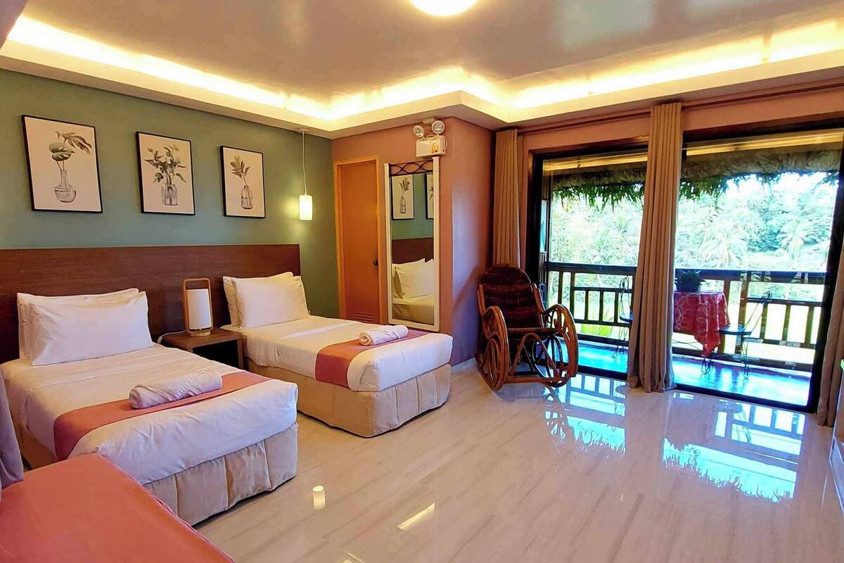 Quarto de hotel com duas camas, uma cadeira de balanço de madeira, varanda com vista para vegetação e quadros nas paredes.