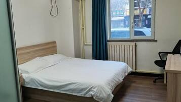 Uma cama de solteiro com cabeceira de madeira, uma janela com cortinas azuis, um radiador e uma escrivaninha com uma cadeira.