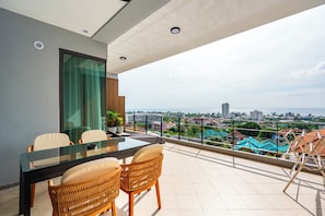 Uma varanda com mesa e cadeiras de vidro, vista para um horizonte urbano e um edifício moderno com telhado inclinado.