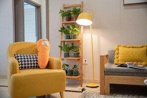 Uma poltrona amarela com almofada estampada, um aparador de madeira para plantas e um abajur com cúpula amarela.