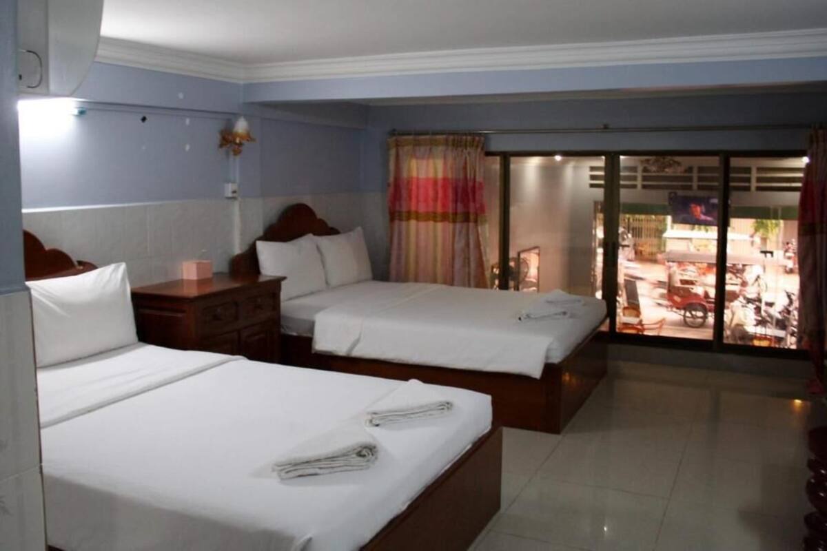 Quarto de hotel com duas camas, uma cômoda de madeira e vista para a rua através de uma porta de vidro.
