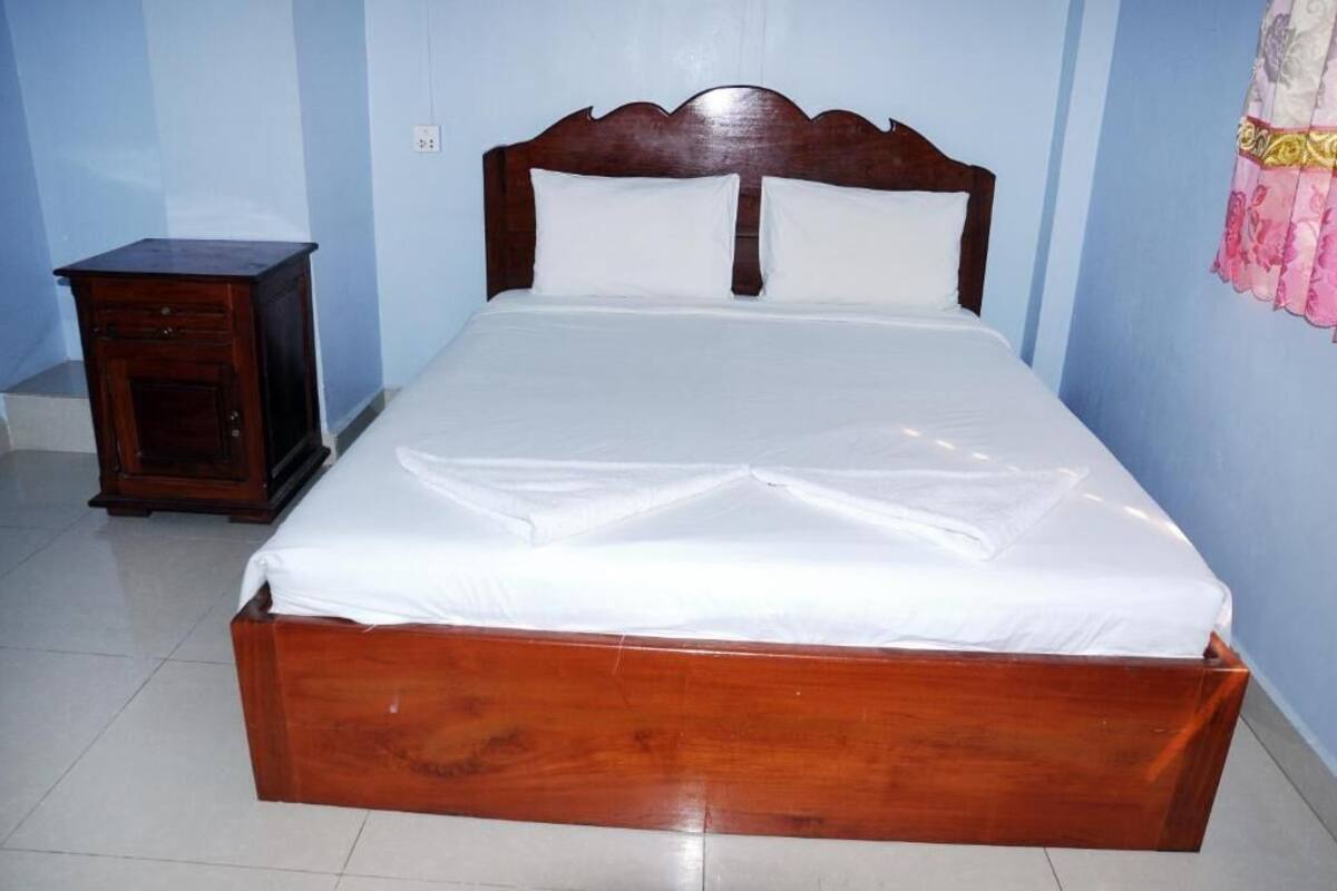 Cama de casal com estrutura de madeira e roupa de cama branca em quarto com paredes azul claro e piso de azulejos.