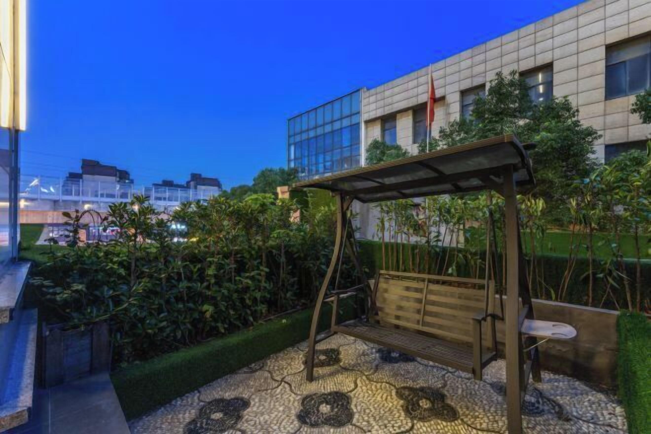 Uma área de estar moderna ao ar livre, com um banco balanço e uma pequena mesa, cercada por vegetação exuberante e um piso estampado.