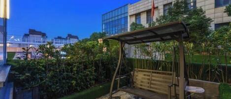 Uma área de estar moderna ao ar livre, com um banco balanço e uma pequena mesa, cercada por vegetação exuberante e um piso estampado.