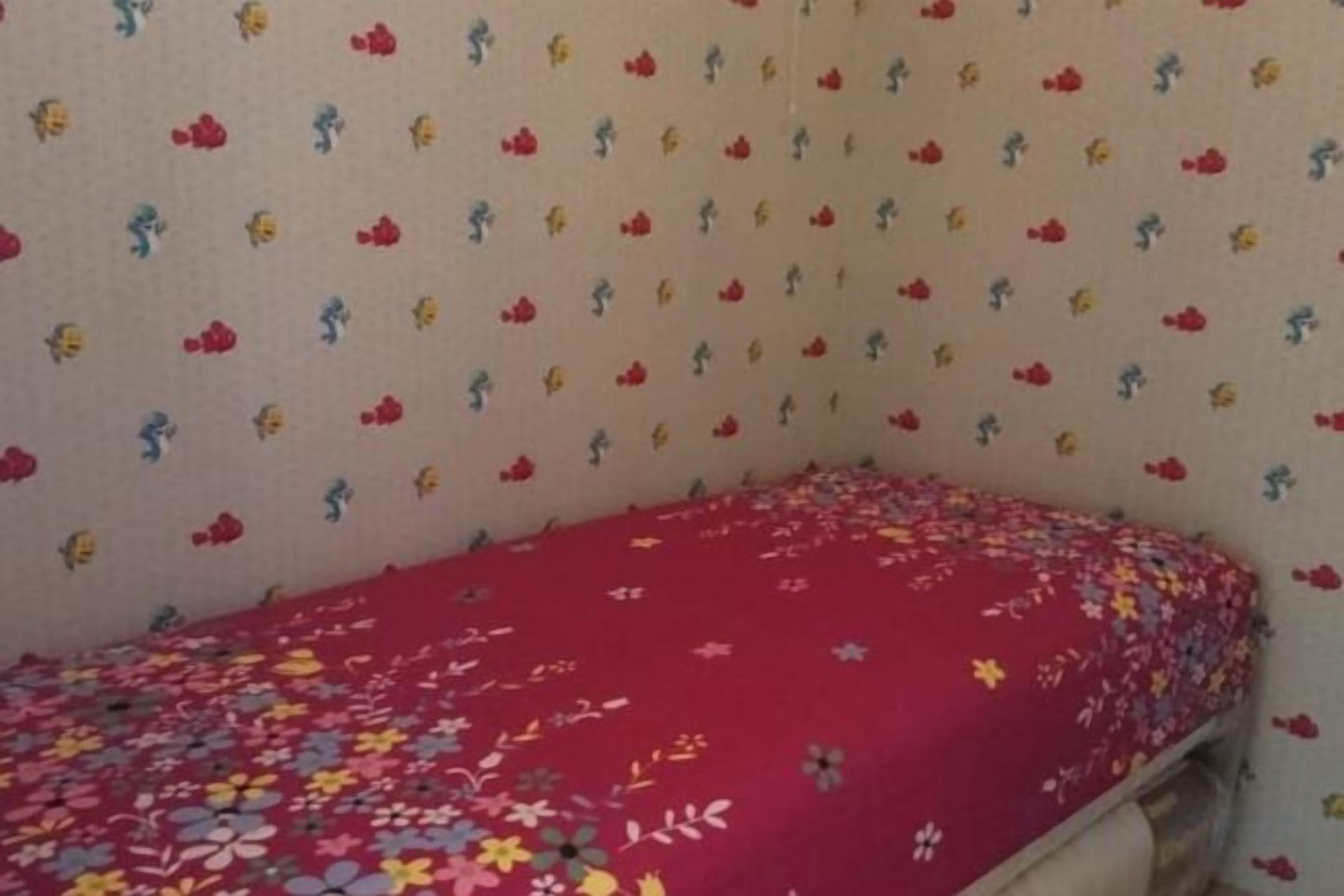 Quarto pequeno com uma cama de solteiro, papel de parede estampado com flores e colcha vermelha.